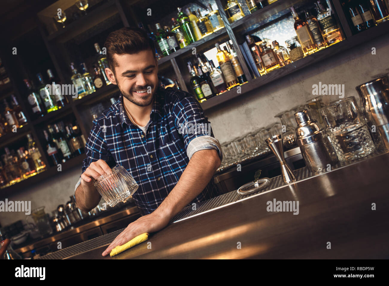 Young barman standing at bar counter holding glasses wiping table ...
