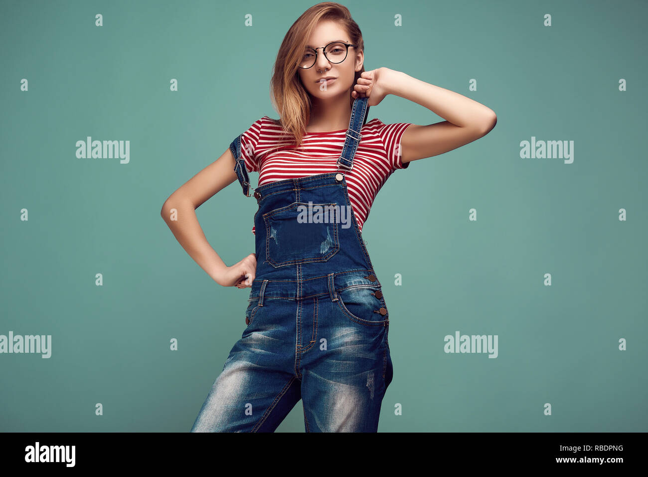 Portrait of cute teenage girl in denim overalls and glasses posing on ...