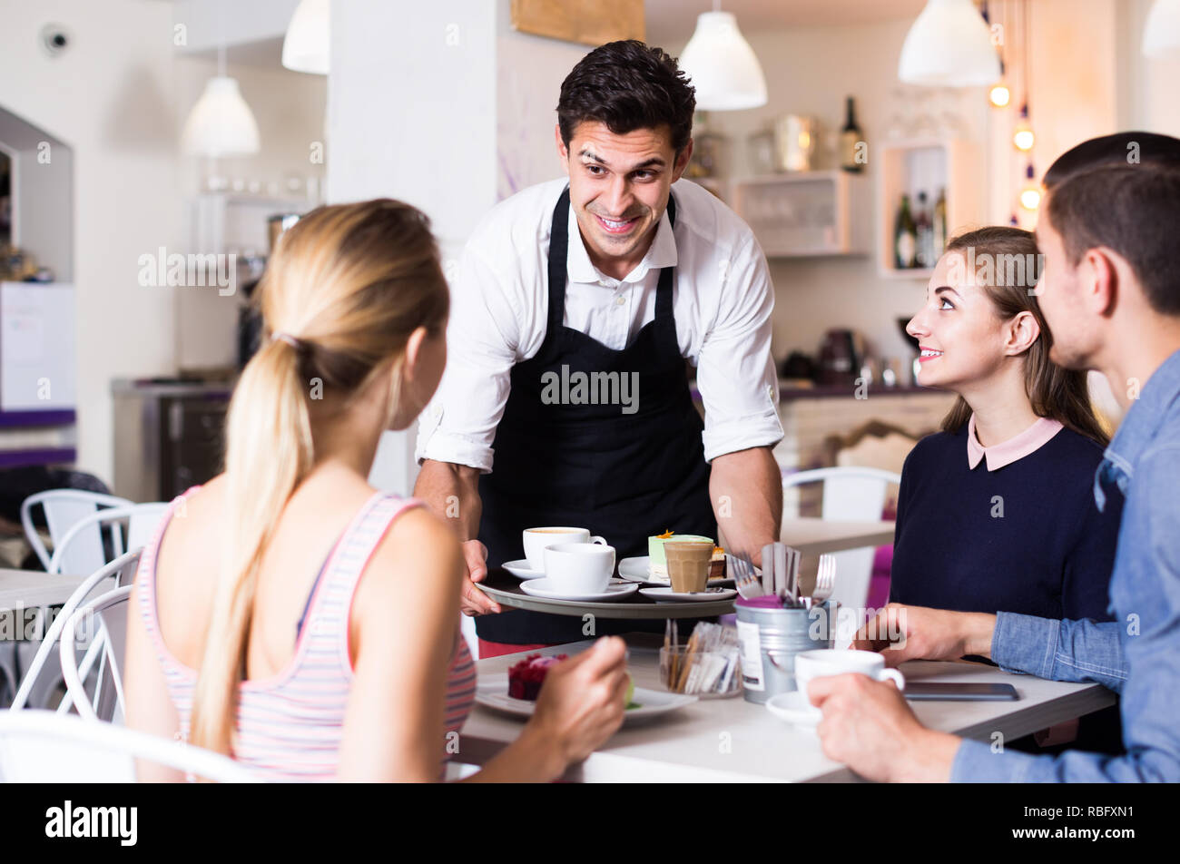 Polite happy waiter bringing ordered dishes to friends in tearoom of a ...