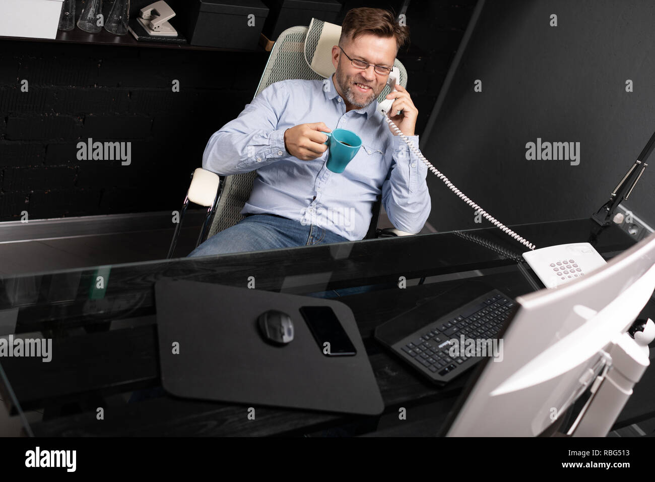 man leaned back in his chair, drinking coffee and talking on landline ...