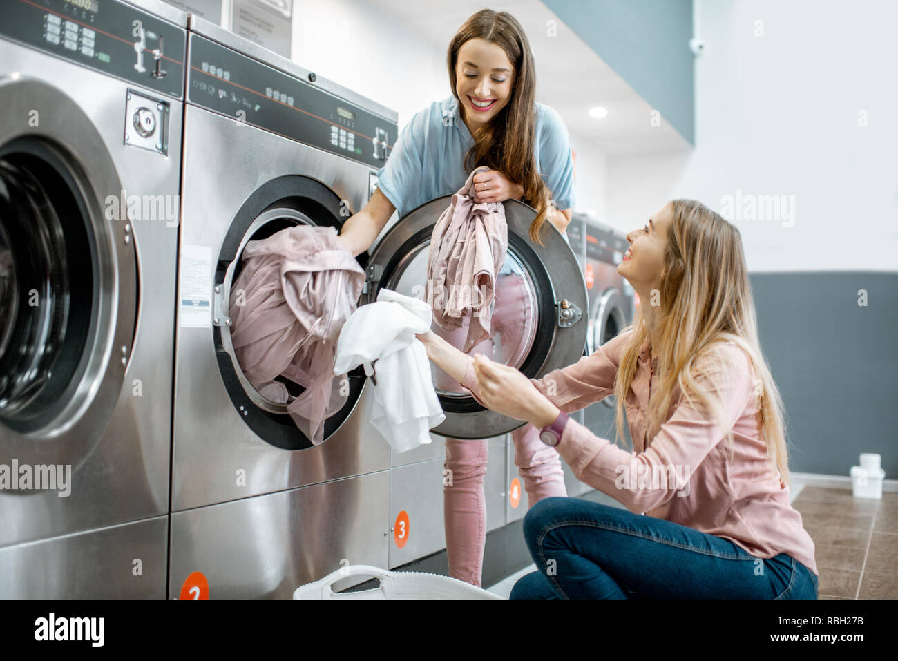 Two cheerful girlfriends having fun loading clothes into the washing ...