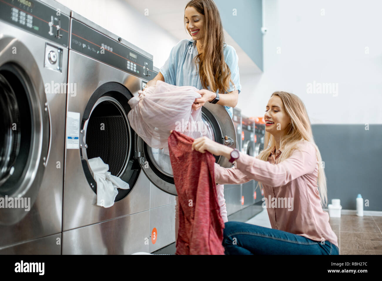 Two cheerful girlfriends having fun loading clothes into the washing ...