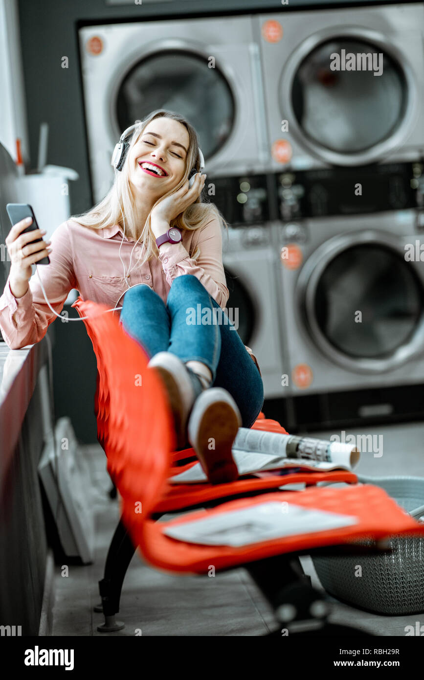 Young Woman Enjoying Music Waiting For The Clothes To Be Washed Sitting On The Chair At The Self