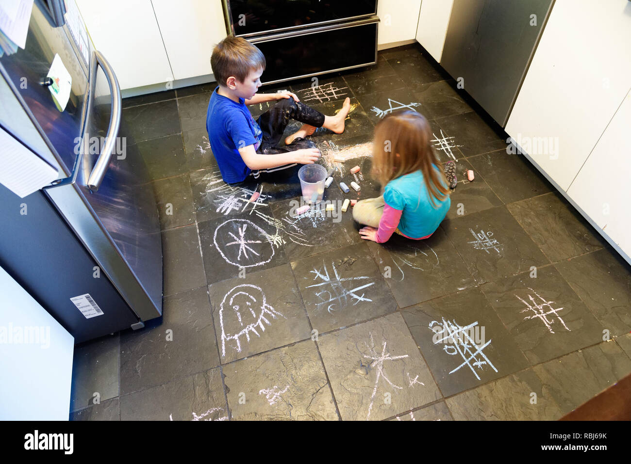 Two children using chalks to draw on a slate floor in their kitchen ...