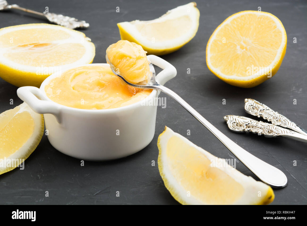 Lemon curd in white jar with spoon with curd and lemon slices over grey ...