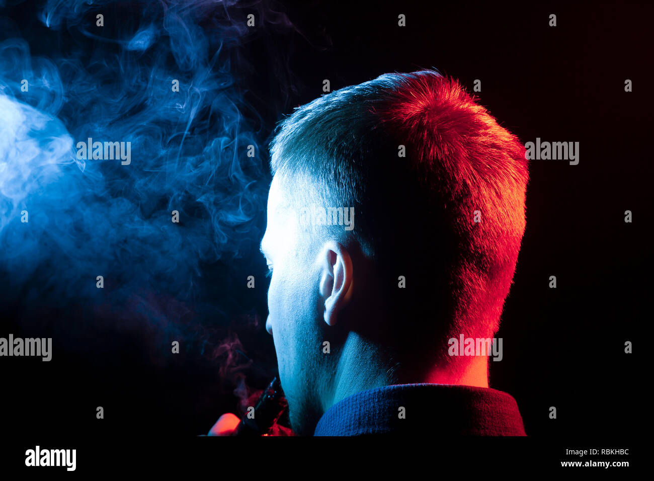 A view from the back of a man’s head around in a shirt smoking a vape ...