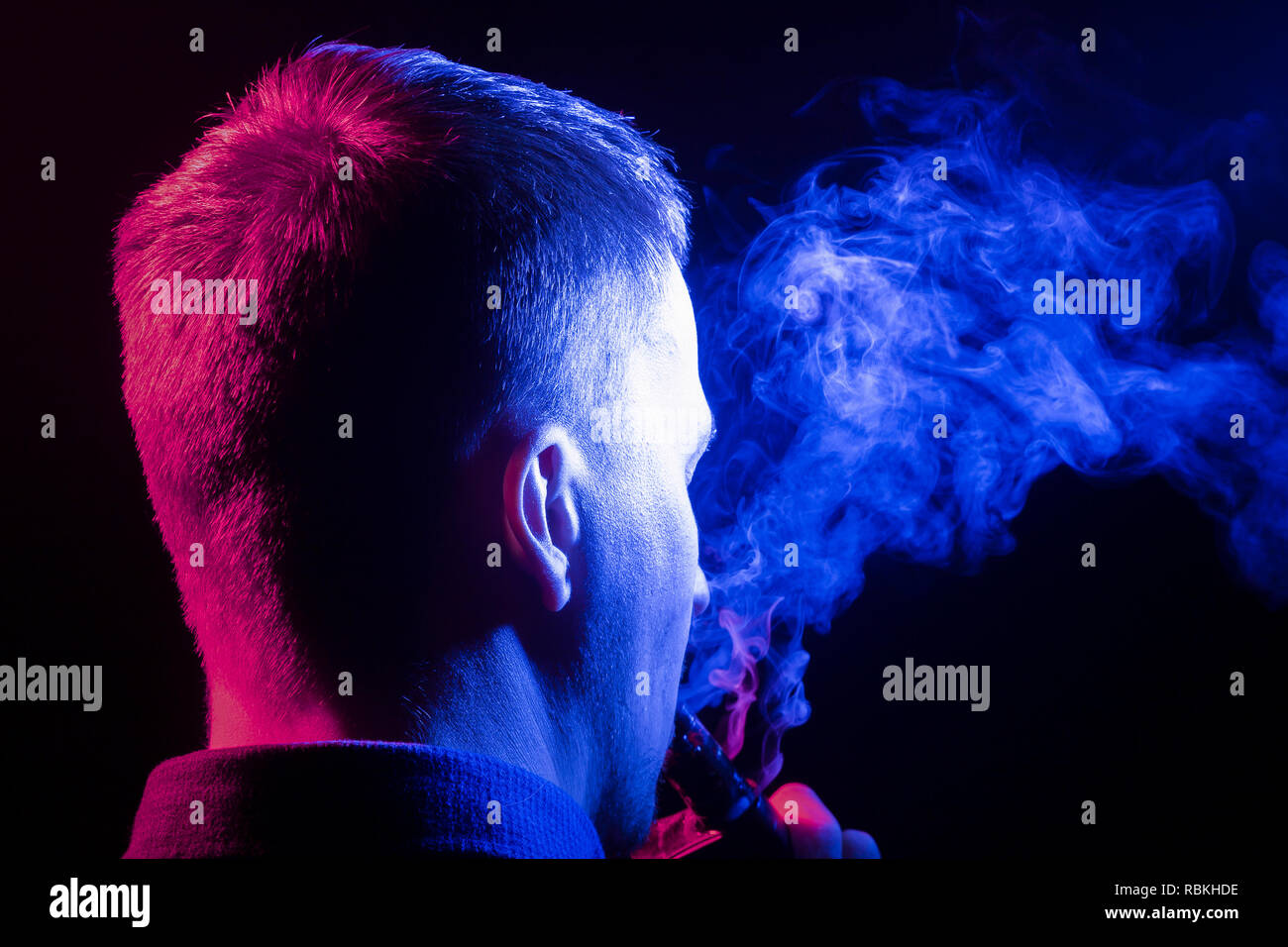 A view from the back of a man’s head around in a shirt smoking a vape ...