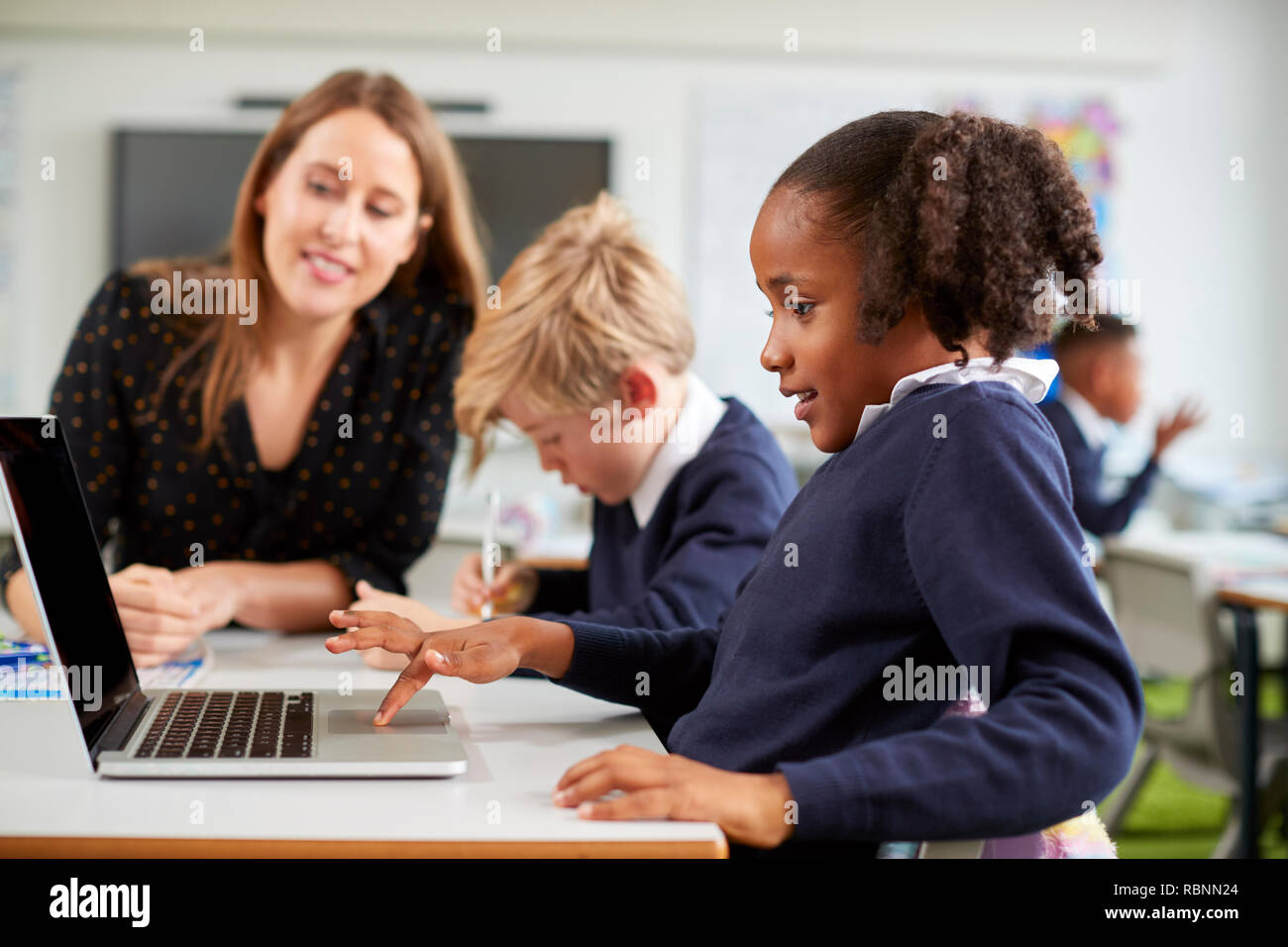 A female school teacher sitting at a desk helping a boy and girl using ...