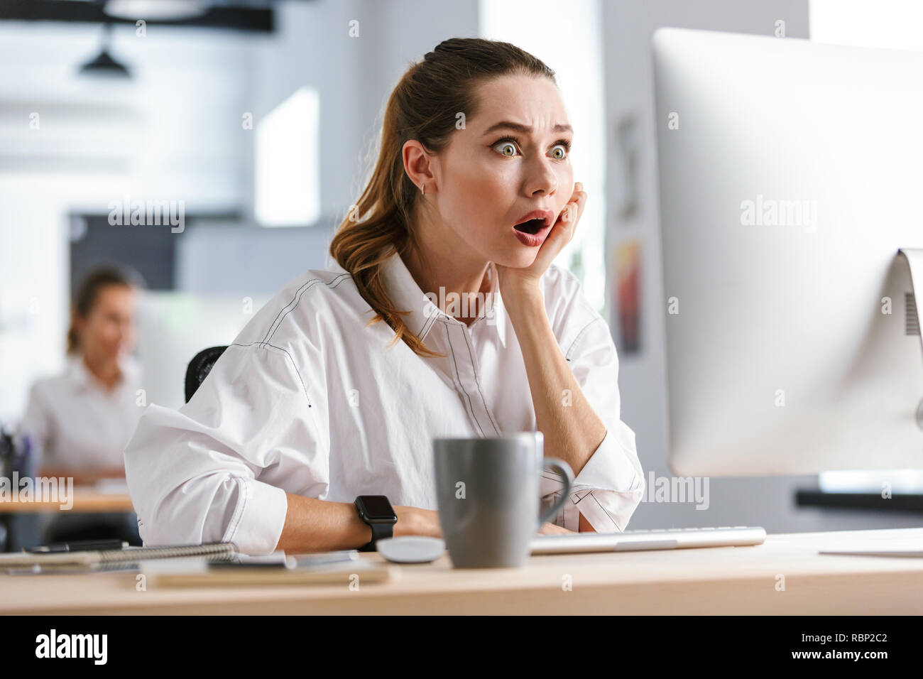 Shocked young woman dressed in shirt sitting at her workplace at the ...