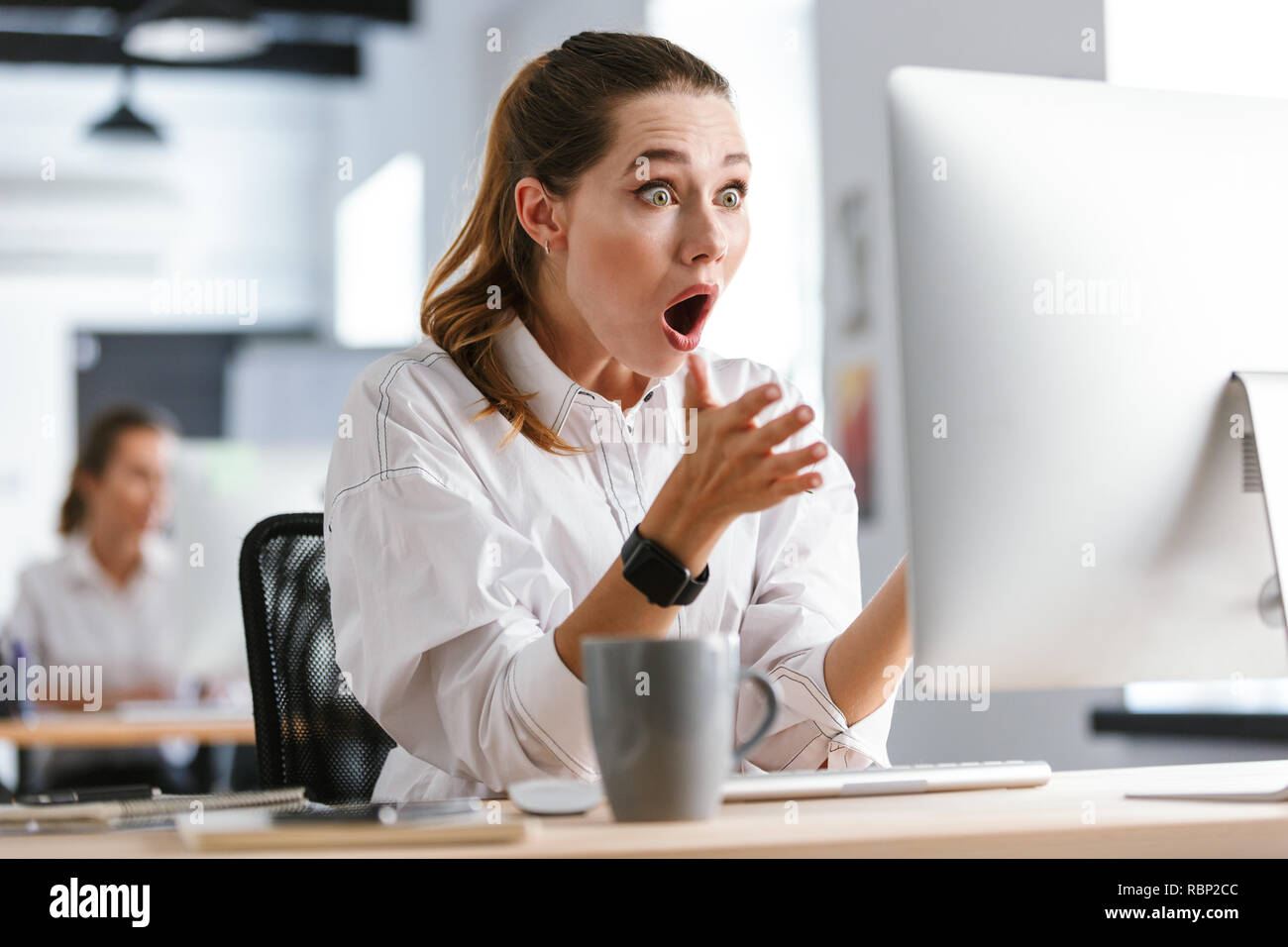 Shocked young woman dressed in shirt sitting at her workplace at the ...