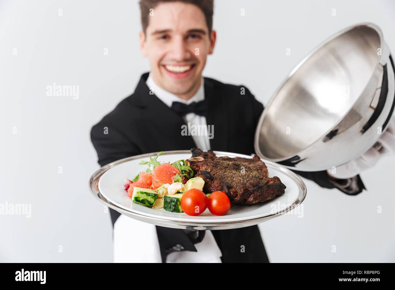 Close up of a happy waiter presenting a meat dish isolated over gray ...