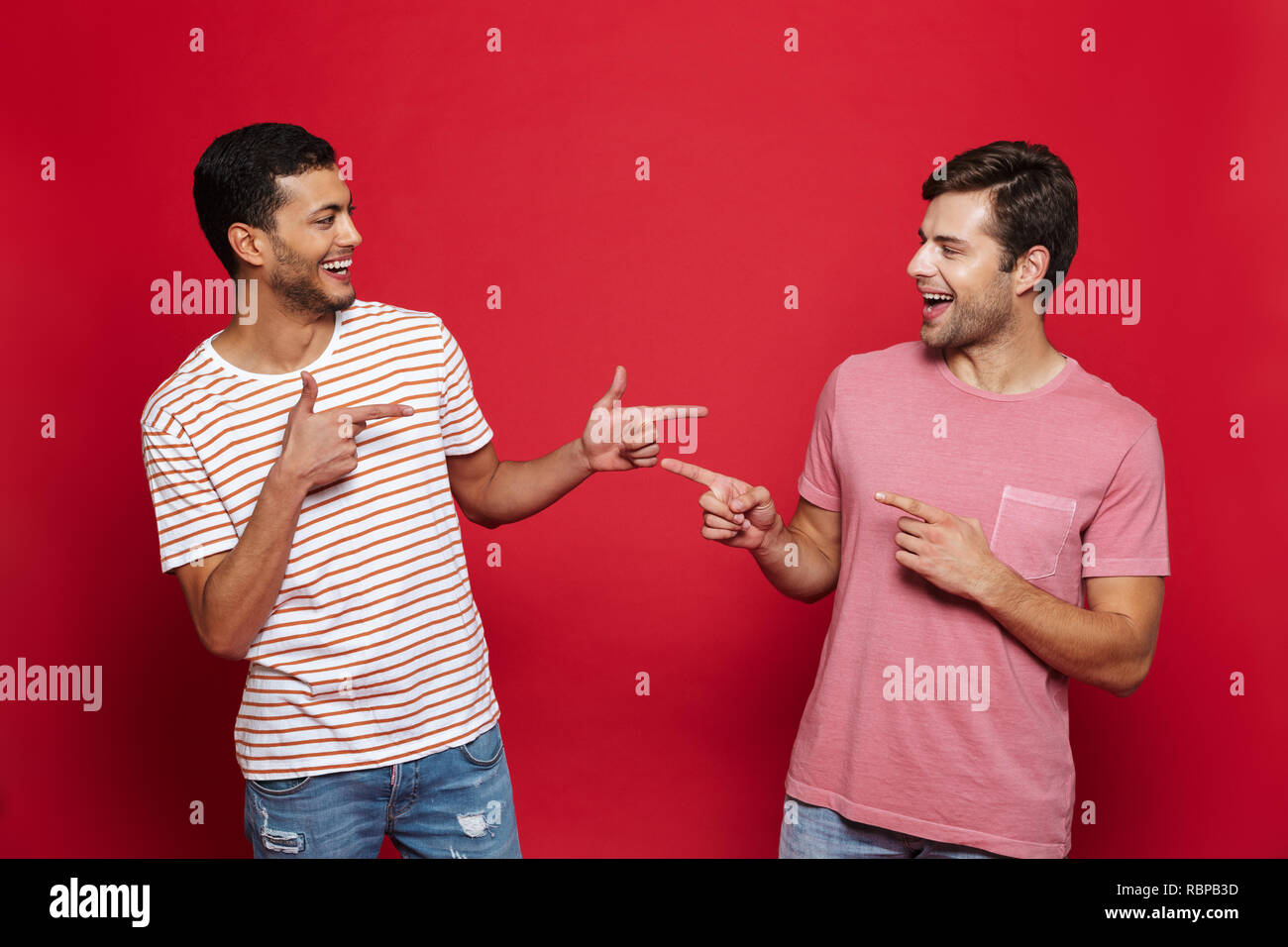 Two cheerful young men standing isolated over red background, pointing ...