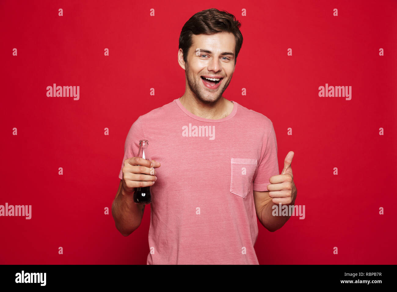 Happy young man holding glass bottle with fizzy drink isolated over red ...