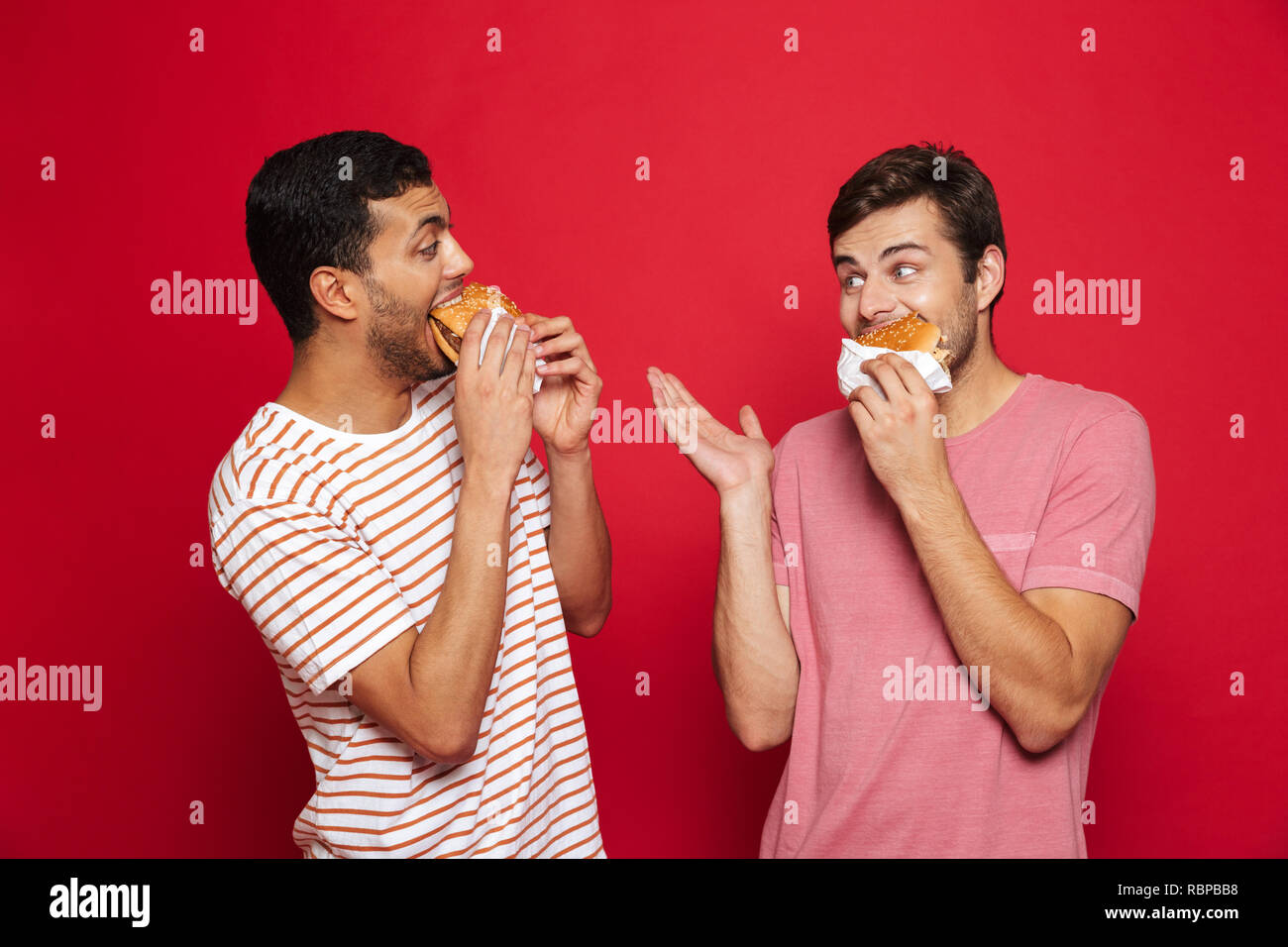 Two cheerful men friends standing isolated over red background, eating ...