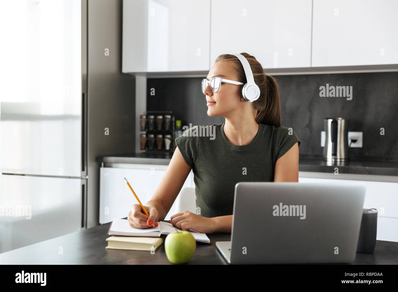 Smiling young woman sitting at the kitchen, using laptop computer for ...