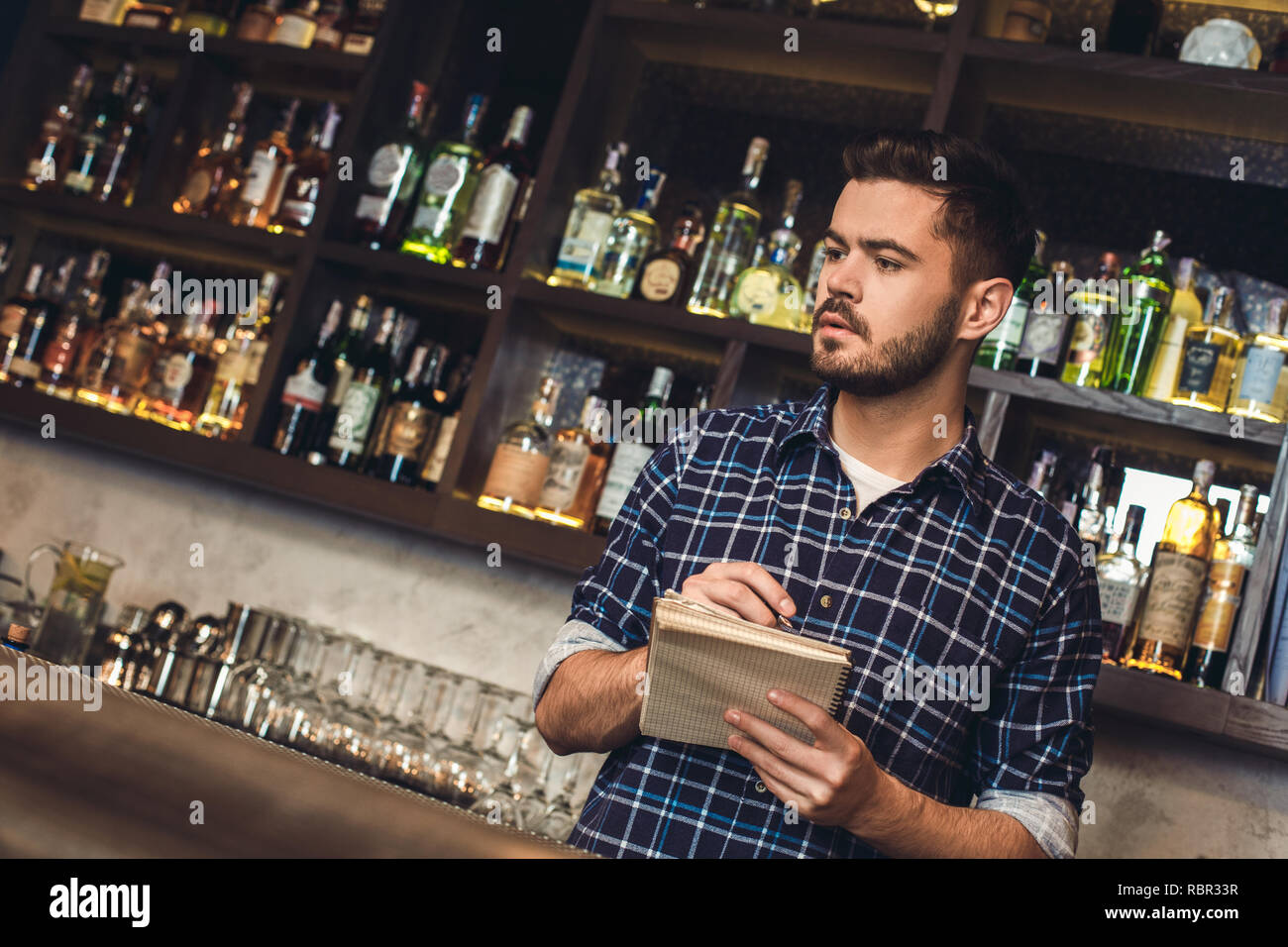 Young barman standing at bar counter checking inventory taking notes ...