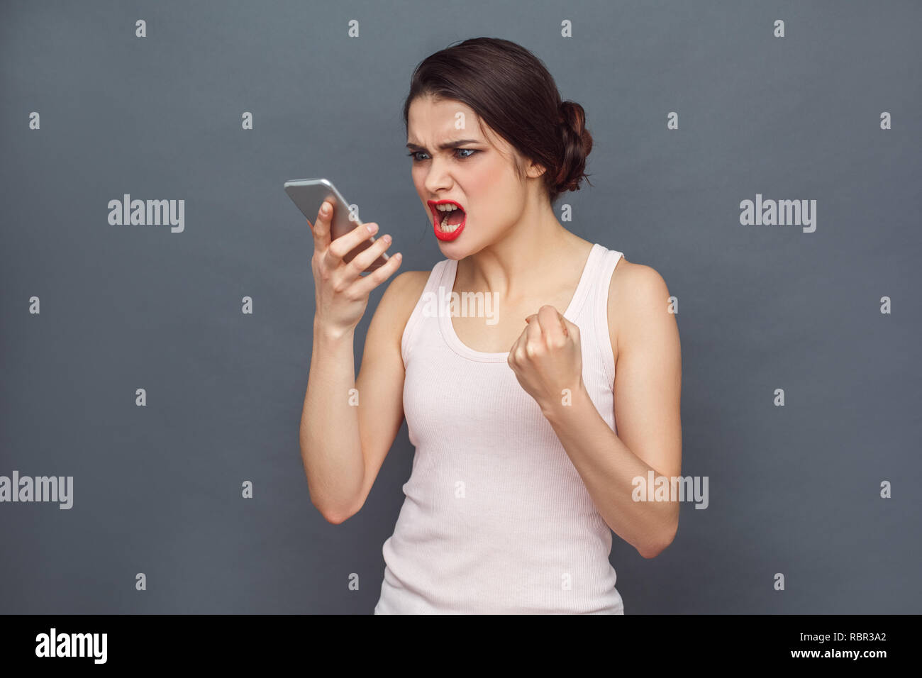 Young woman standing isolated on grey wall holding smartphone shouting ...