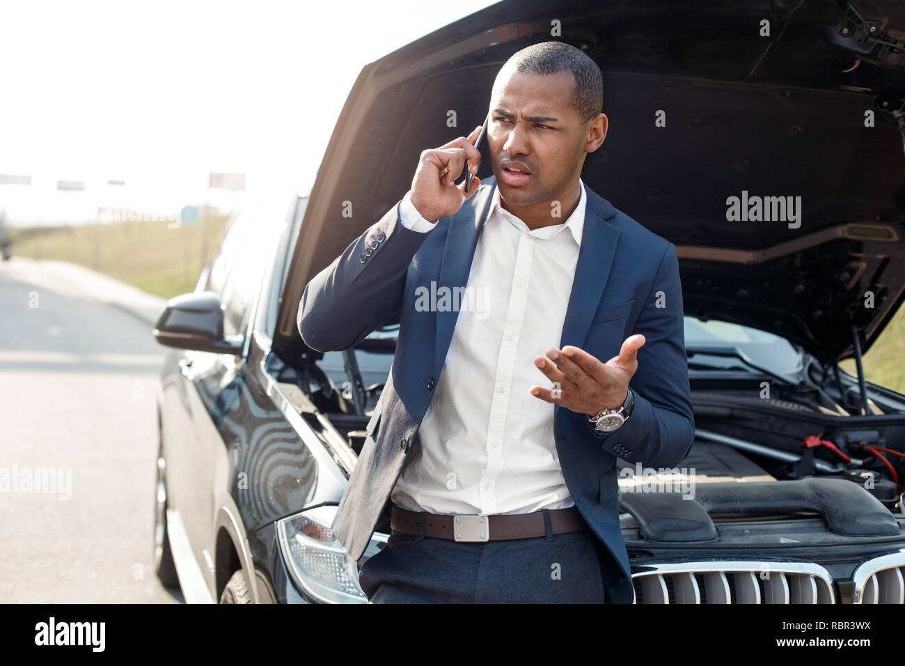 Young mulatto man leaning on car with opened hood talking on smartphone ...