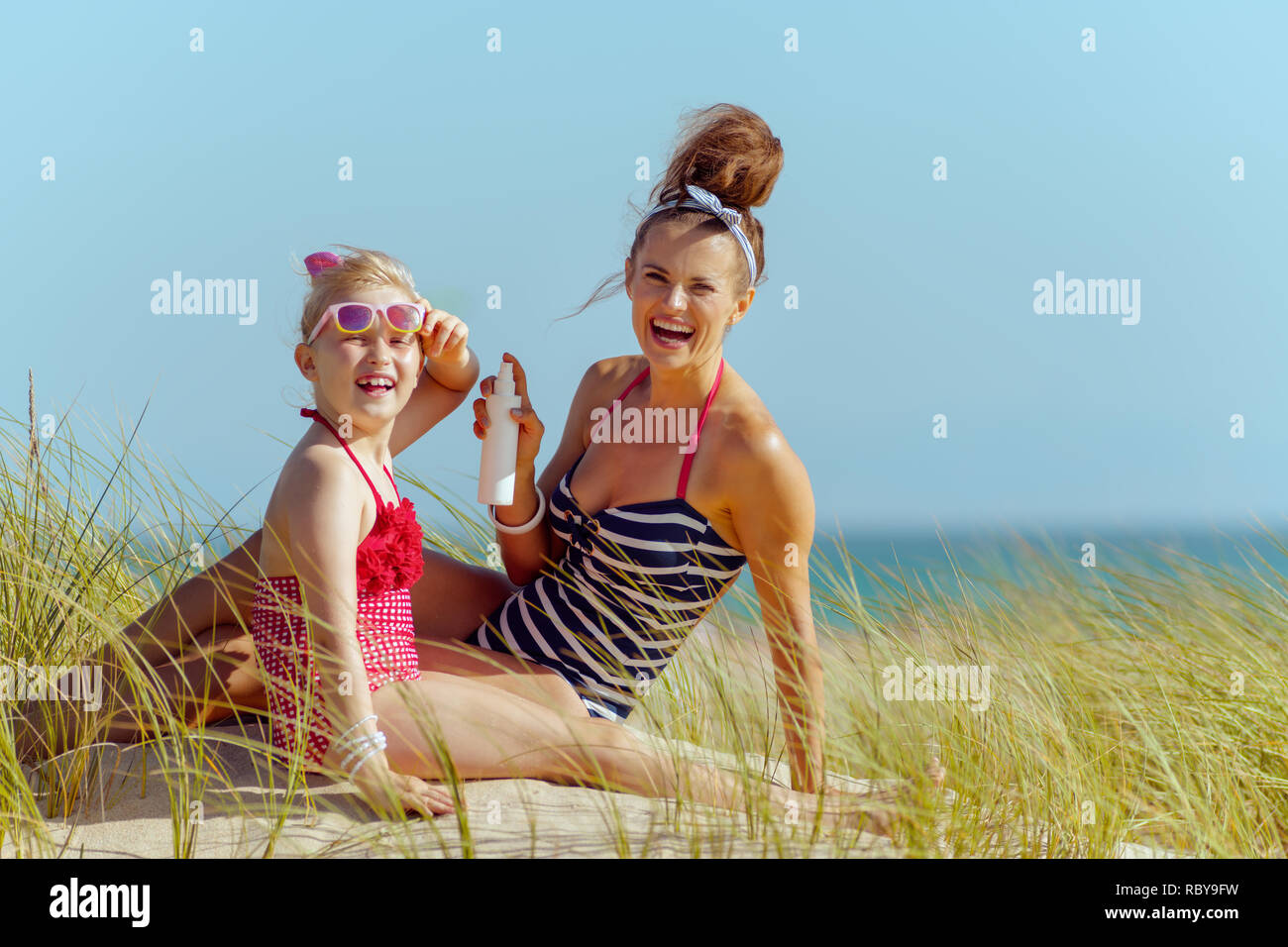 Smiling Young Mother And Child In Beachwear On The Seashore Applying Suntan Lotion Sunscreen smiling-young-mother-and-child-in-beachwear-on-the-seashore-applying-suntan-lotion-sunscreen