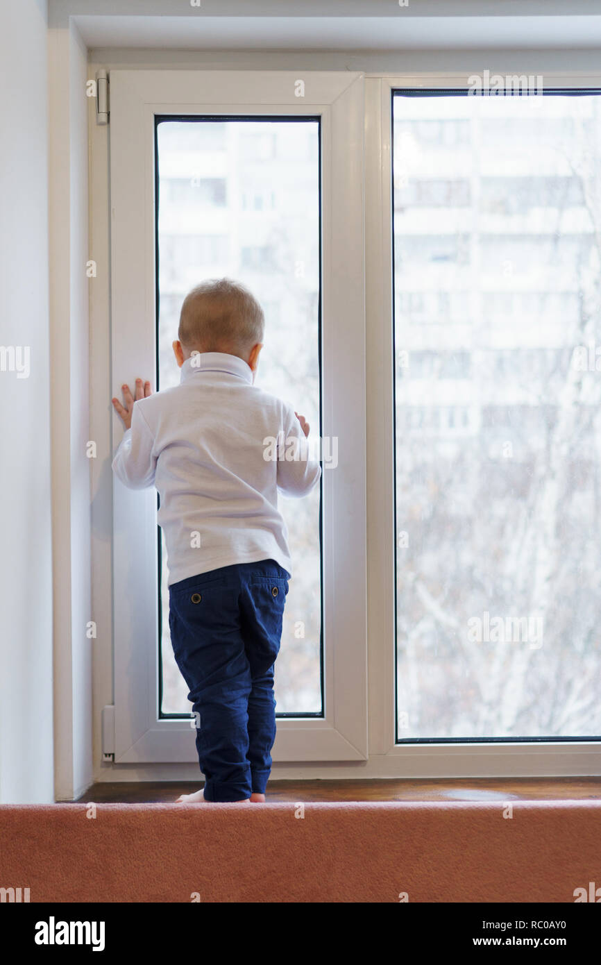 Little boy stands on windowsill and looks out window. Child is ...