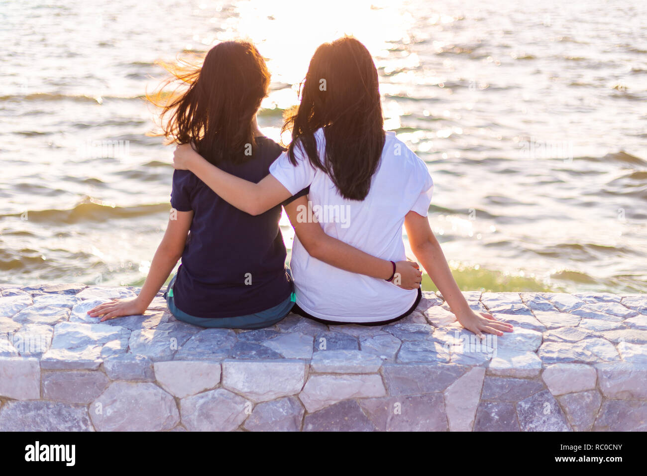 Two young sisters sitting on stone wall watching sunset over the ocean ...