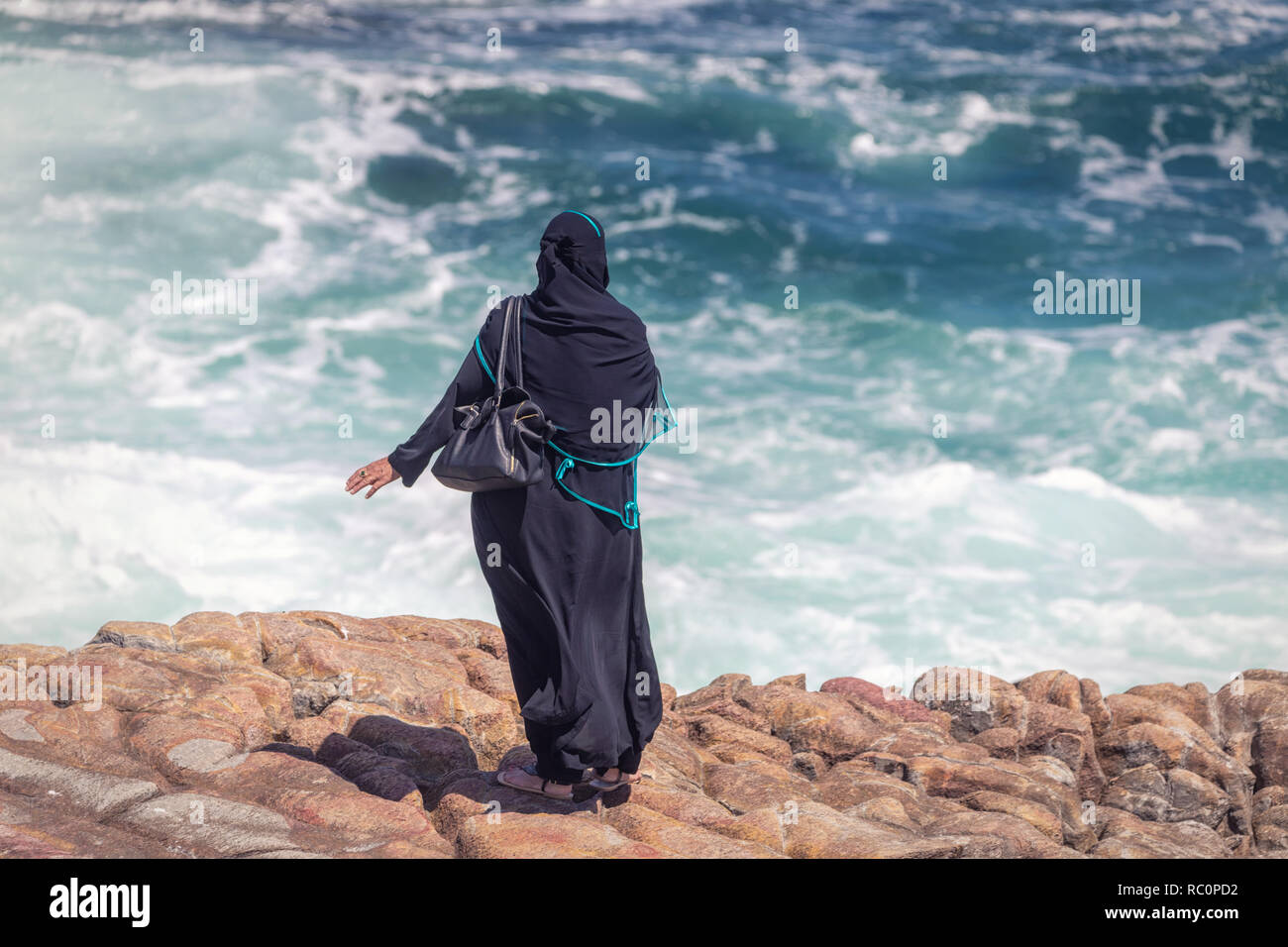 Arabic woman in traditional clothes looking at big waves in Hermanus ...