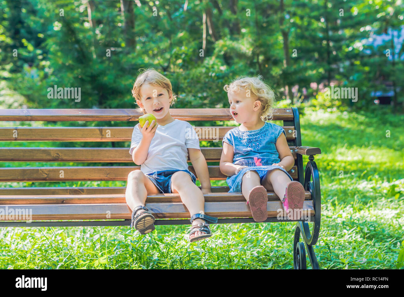 Toddlers boy and girl sitting on a bench by the sea and eat an apple ...