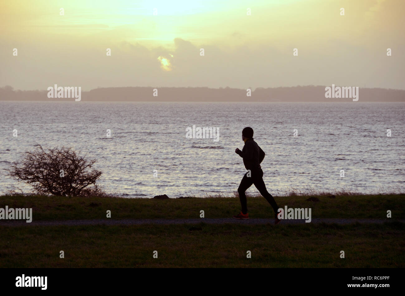 Silhouette of a person jogging in evening light, with the sea as ...