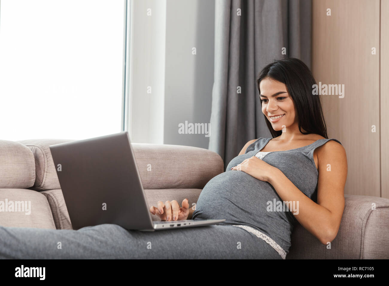 Beautiful young pregnant woman at home, sitting on a couch, using ...