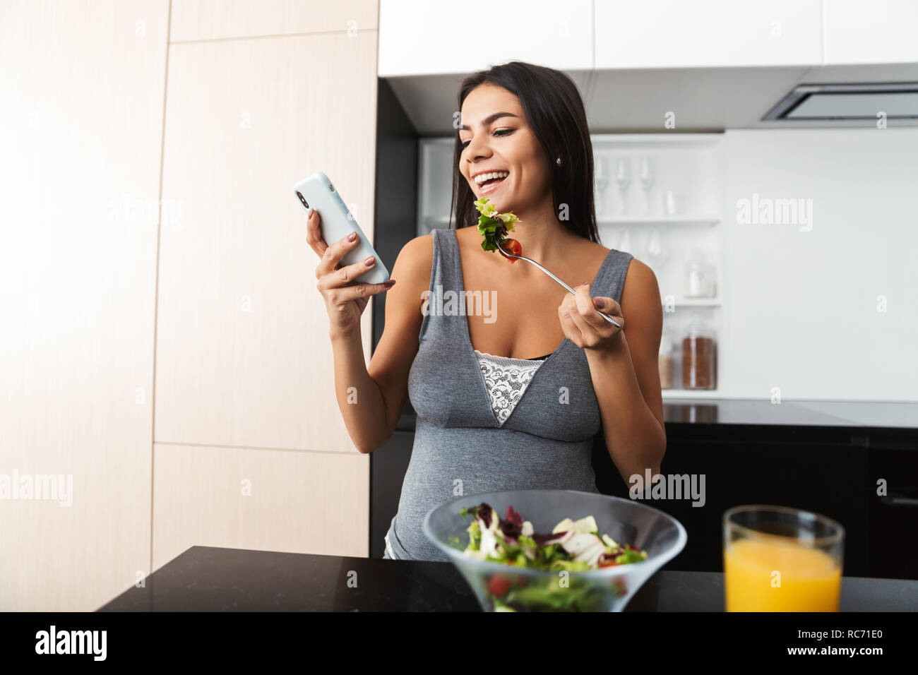 Image of an amazing healthy pregnant woman indoors at home at the kitchen using mobile phone eat ...