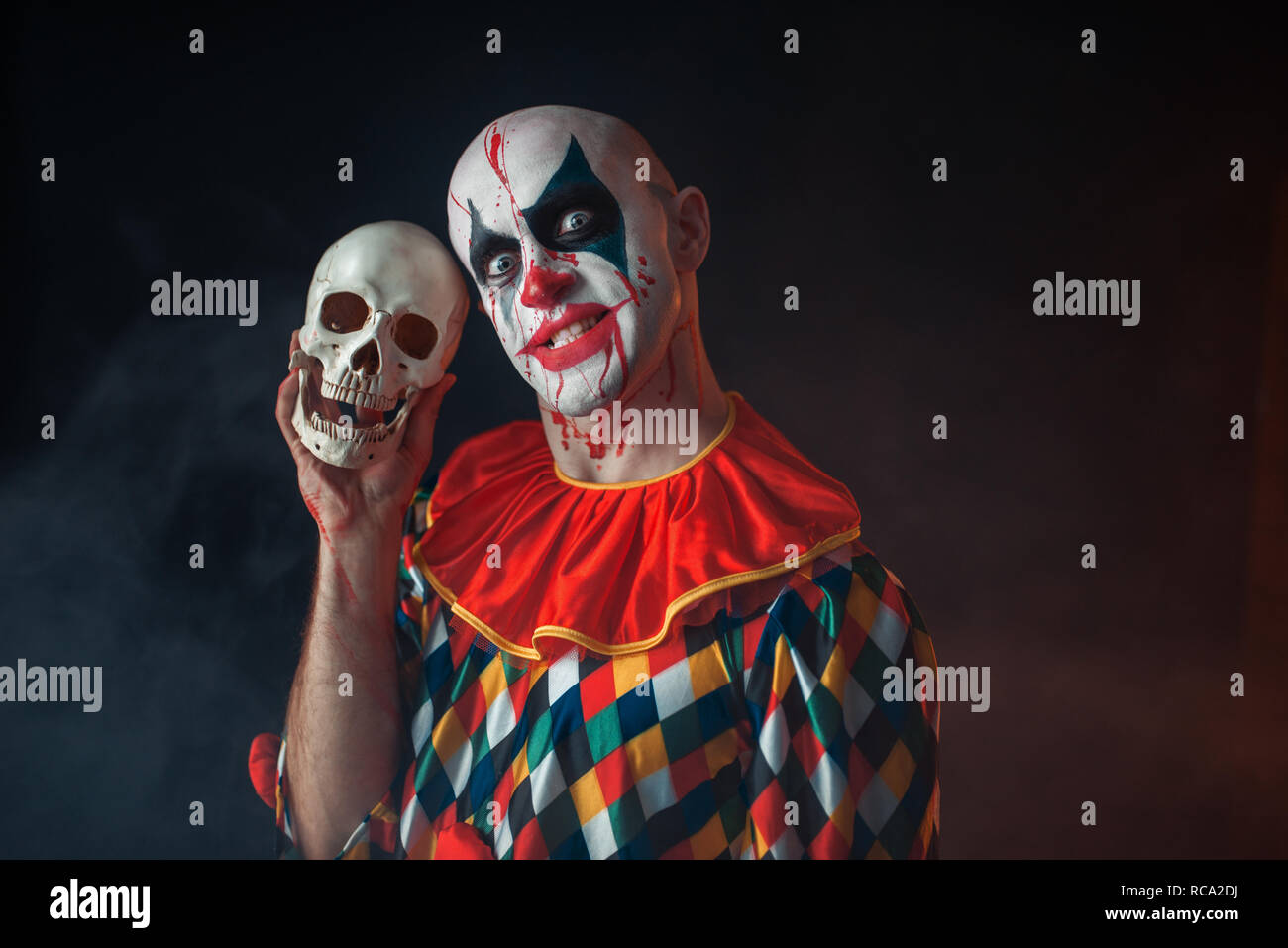Bloody clown with crazy face holds human skull, terrible secret. Man ...