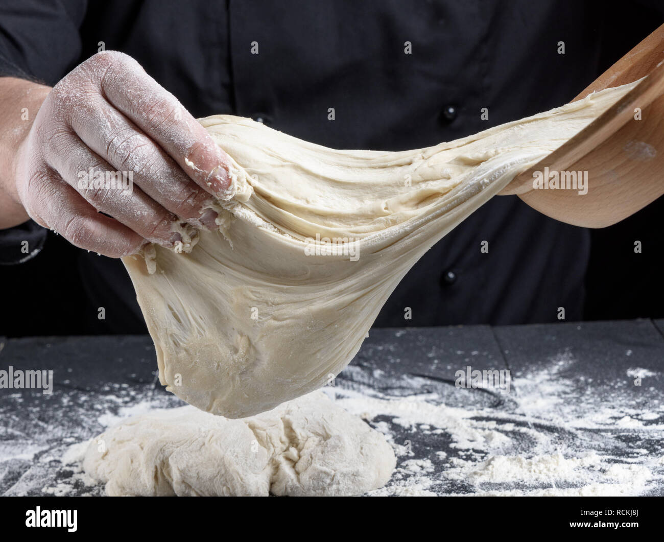 cook in black uniform kneads white wheat flour dough on a wooden table ...