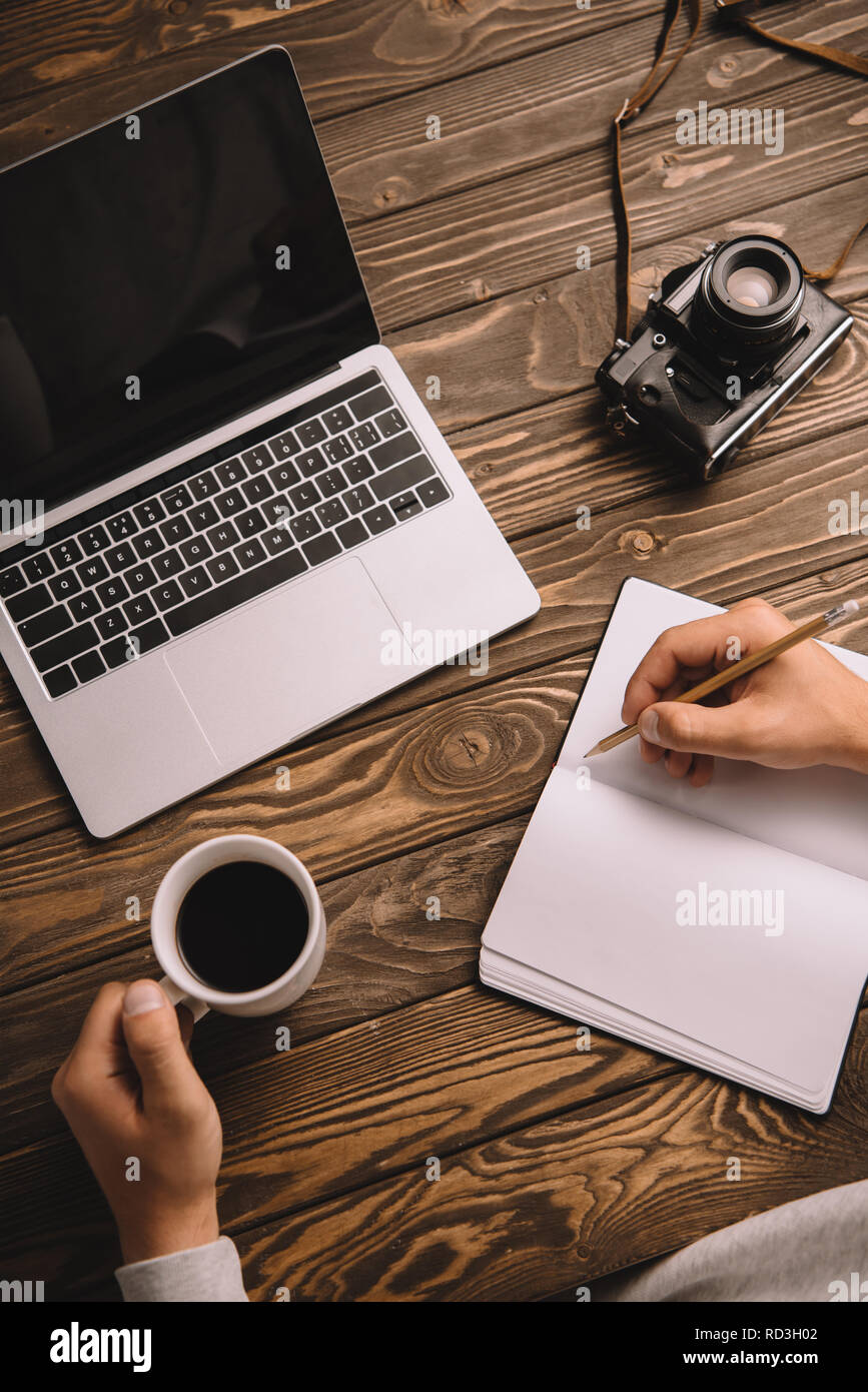 cropped view of male photographer writing in notebook at table with cup ...