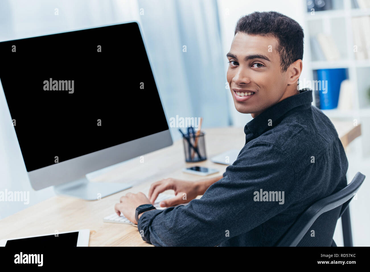 young mixed race businessman using desktop computer with blank screen and smiling at camera ...
