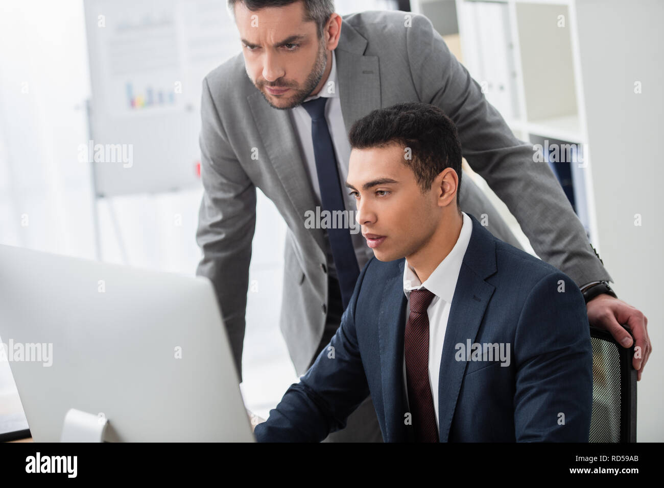 serious business mentor looking at young colleague using desktop ...