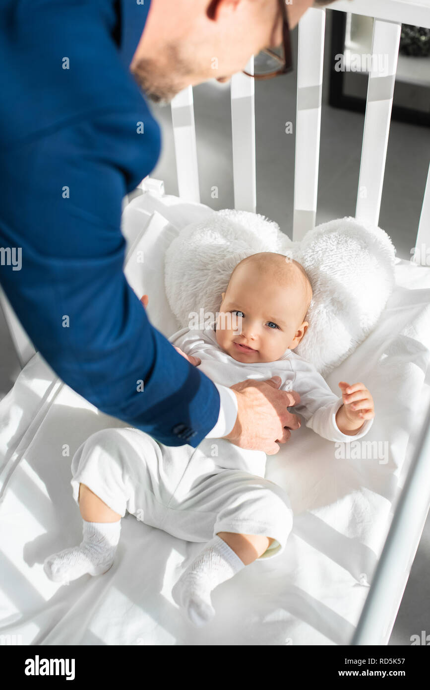 cropped view of father in suit putting infant daughter into baby crib ...