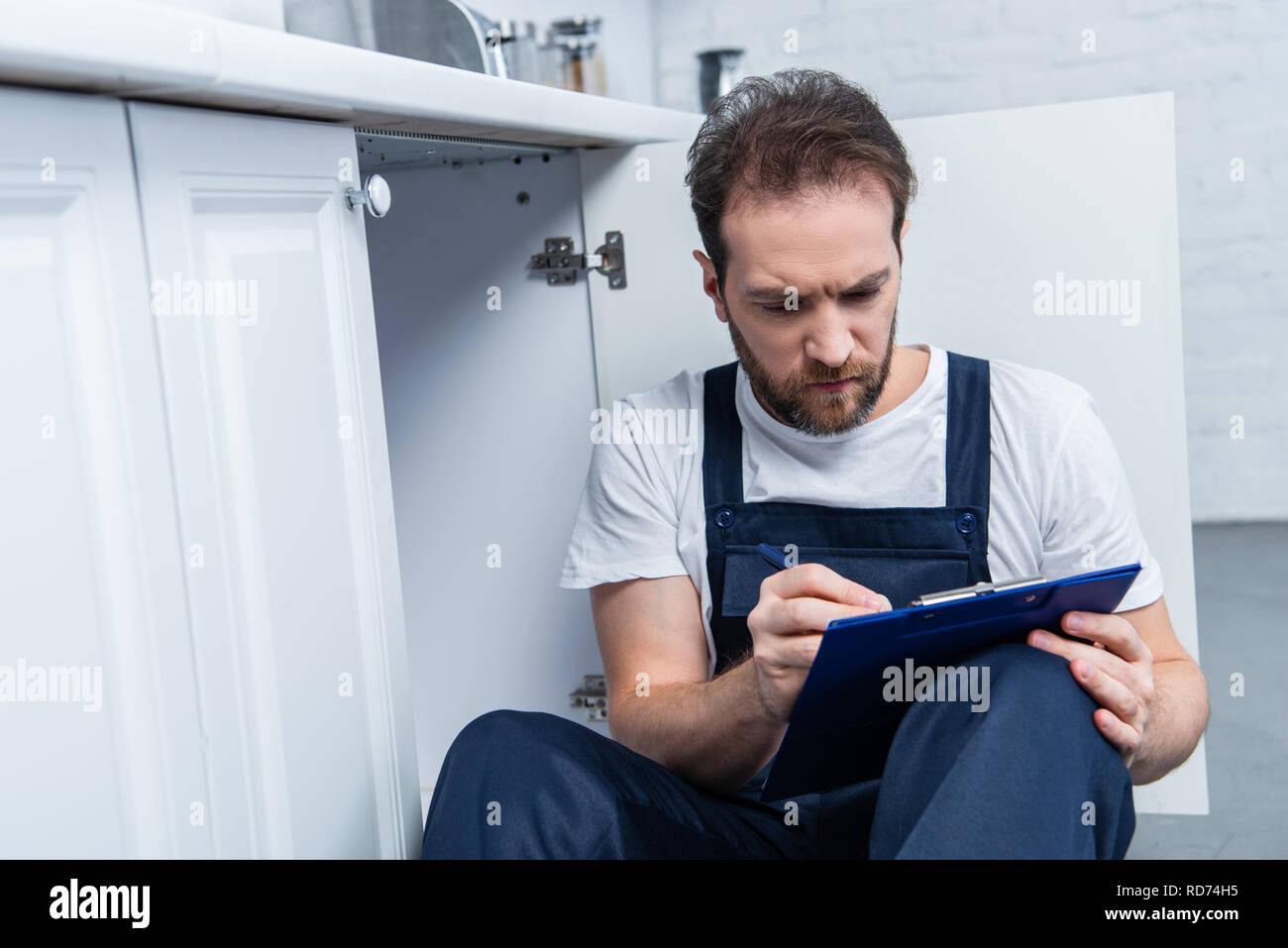 serious adult craftsman writing in clipboard while sitting on floor in ...