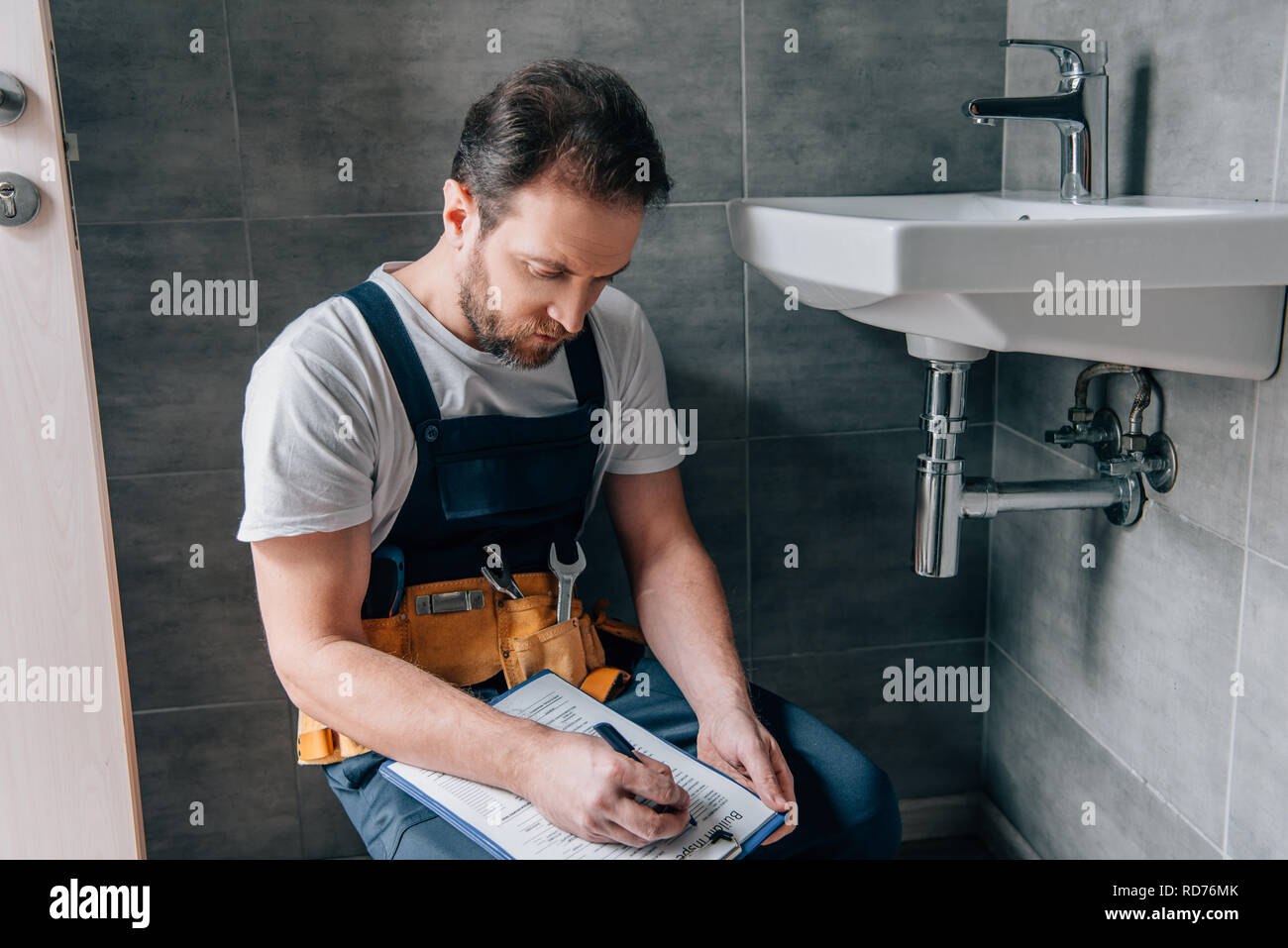 adult male plumber with toolbelt writing in clipboard near broken sink ...