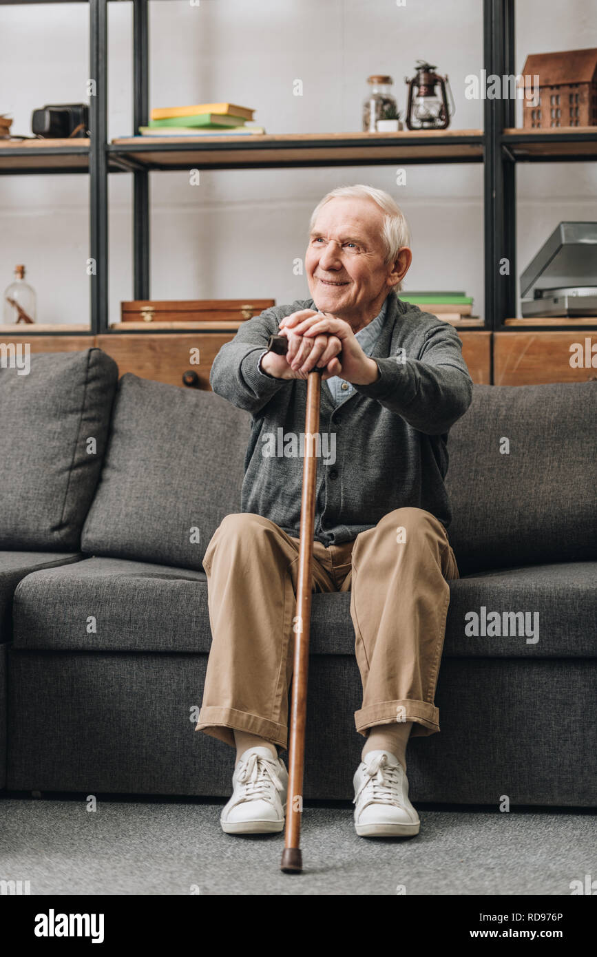 cheerful pensioner smiling and holding walking stick and sitting on ...
