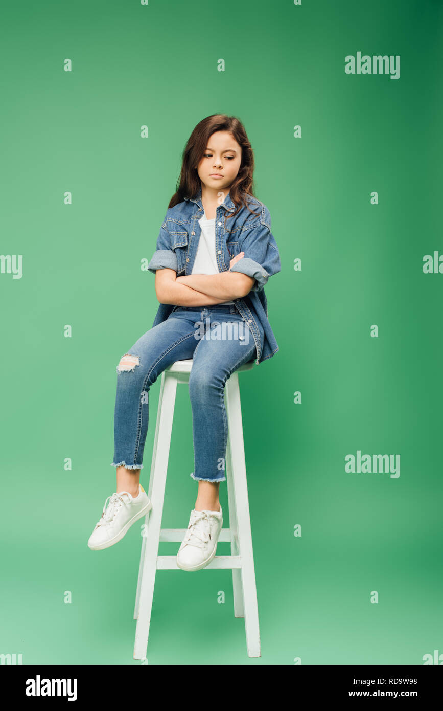 sad child in denim sitting on chair with arms crossed on green ...