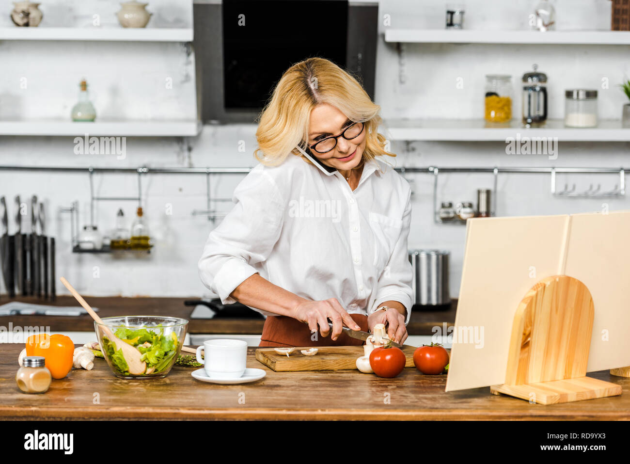 attractive mature woman talking by smartphone, cutting vegetables and ...