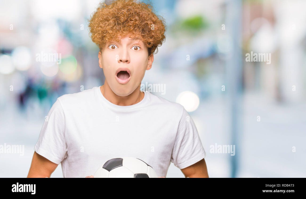 Young handsome man holding soccer football ball scared in shock with a ...