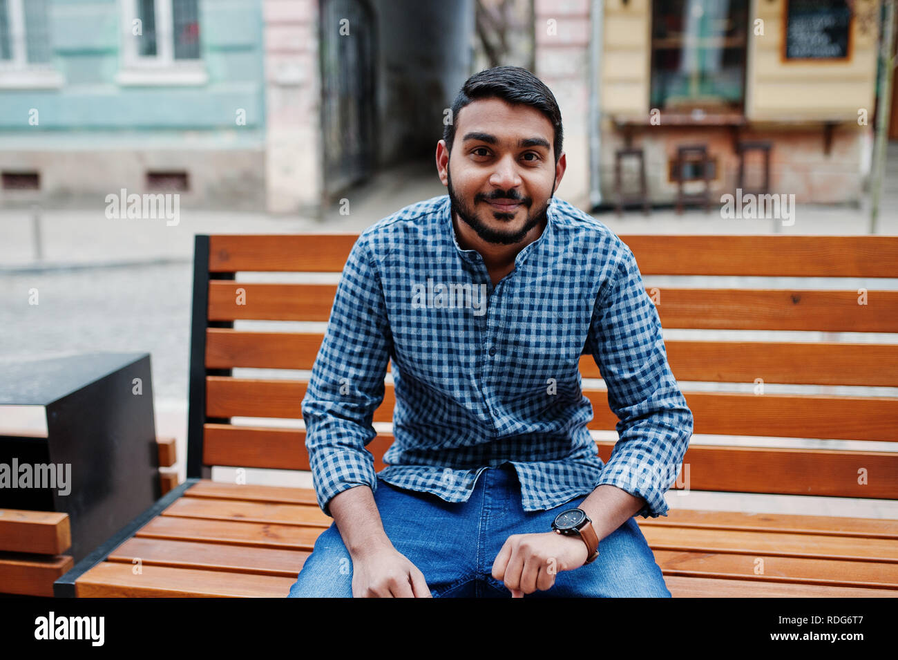 Stylish indian beard model man in casual clothes posed outdoor, sitting on bench at street of ...