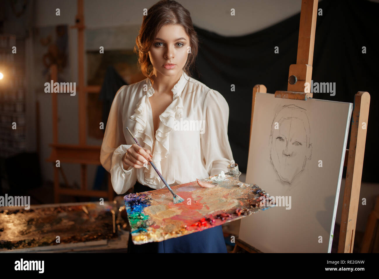 Female painter with color palette and brush standing against easel in ...