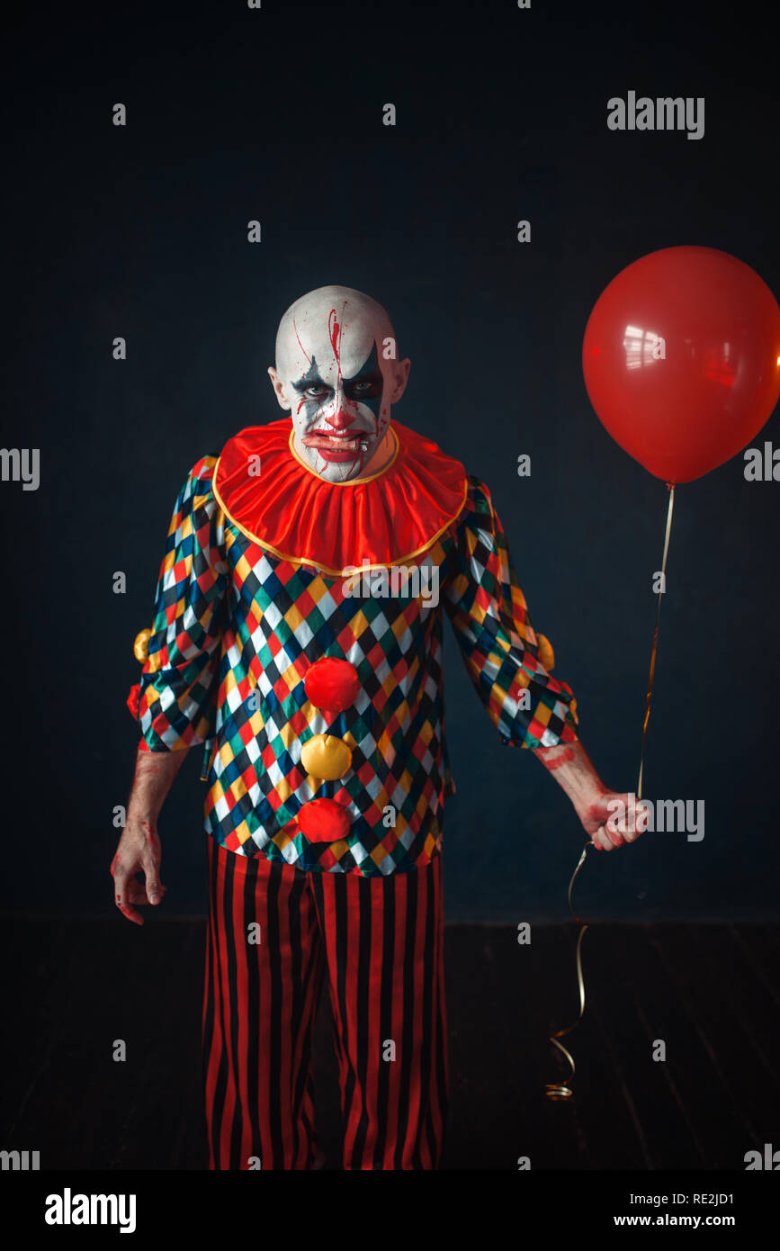 Ugly bloody clown with human finger in his teeth holds air balloon ...