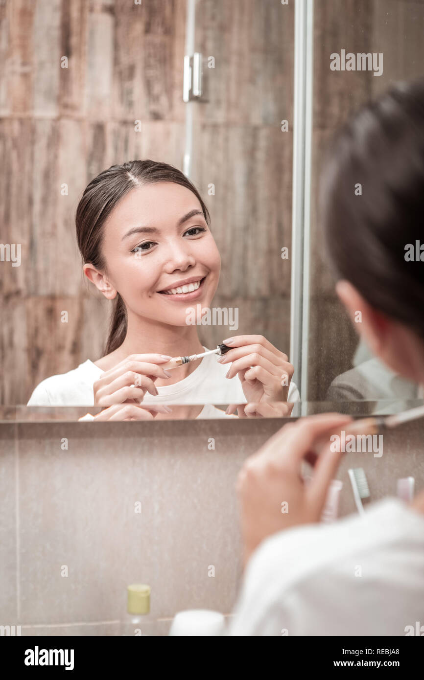 Dark-eyed woman smiling while looking into mirror and doing makeup ...