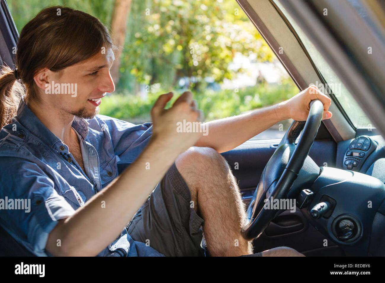 Young man wearing white shirt having long hair, driving car smiling and ...