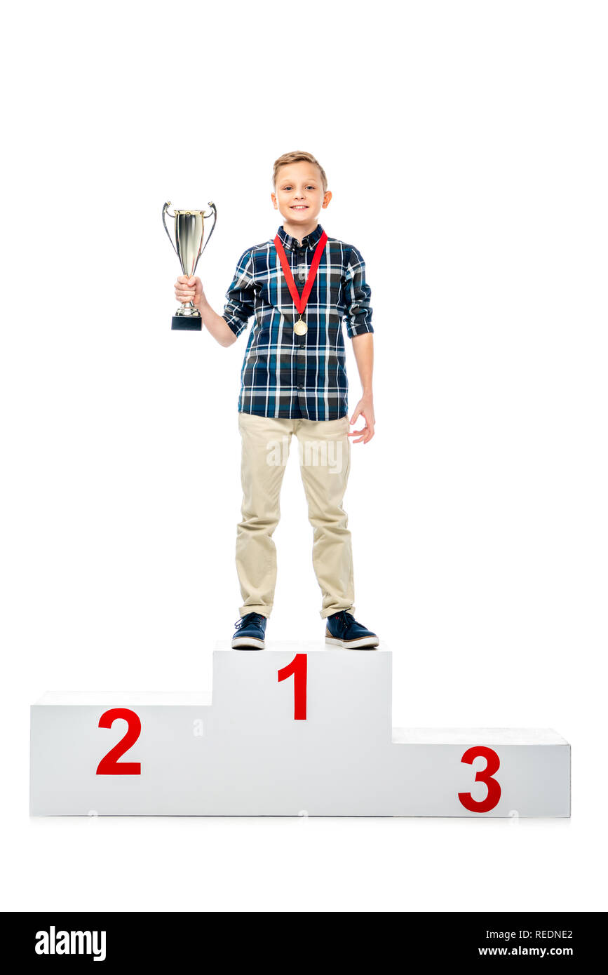 smiling boy standing on winner pedestal, holding trophy cup and looking ...