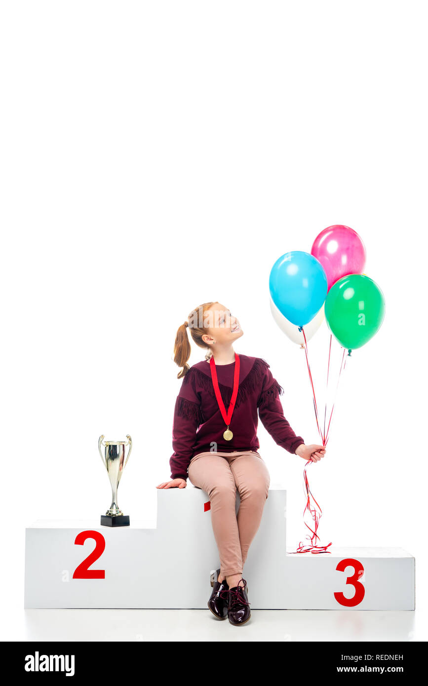 smiling schoolgirl sitting on winner podium near trophy cup and holding ...