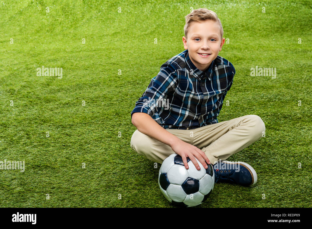 smiling boy sitting on lawn with hand on soccer ball and looking at ...